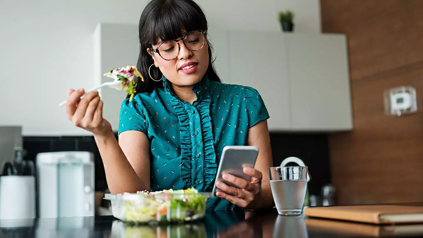 jonge Spaanse Latijns-vrouw die salade eet en naar de telefoon kijkt