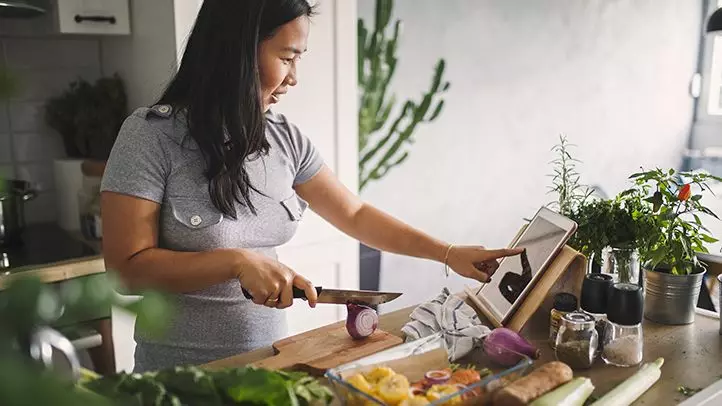 Babaeng Asyano na naghihiwa ng gulay na tumitingin sa tablet sa kusina